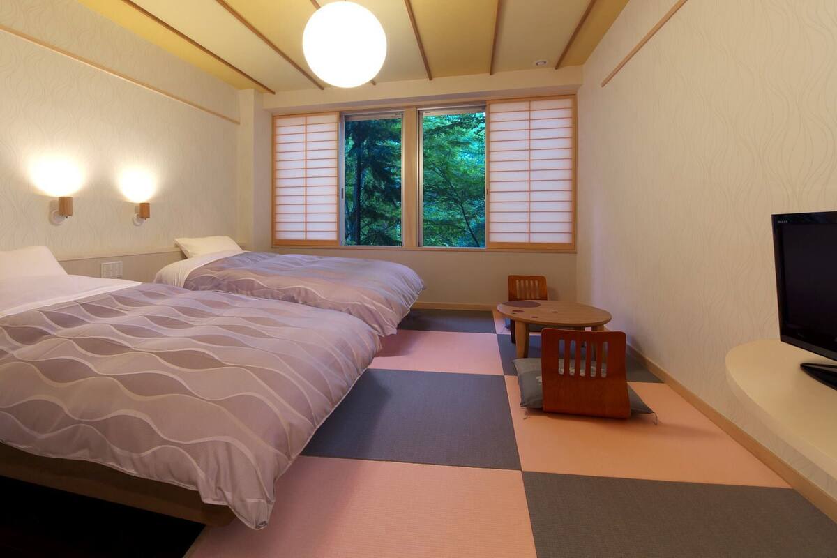 Quarto com duas camas, uma mesa pequena, televisão e janela com painéis shoji.