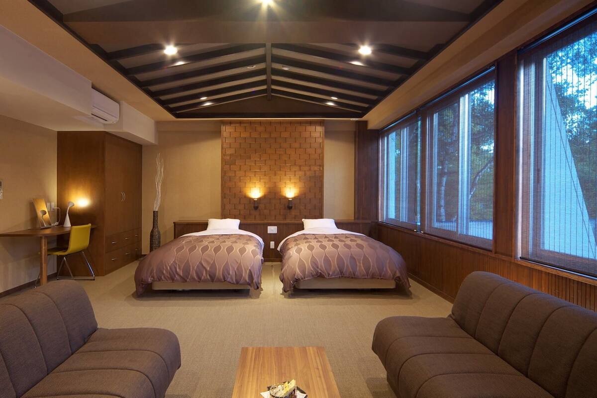 Quarto de hotel com duas camas, uma escrivaninha, uma cadeira e janelas amplas.