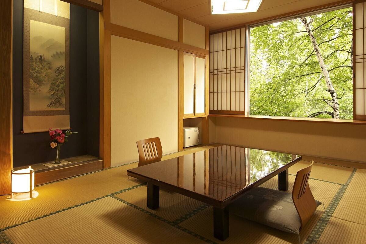 Um quarto japonês tradicional com tatames, uma mesa baixa, uma cadeira, um painel shoji e vista para o verde lá fora.