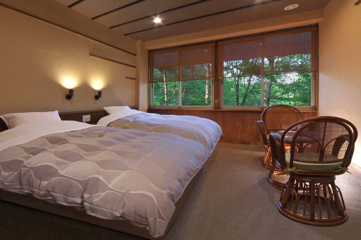 Quarto com uma cama grande, duas luminárias de cabeceira, uma janela com vista para árvores e uma cadeira de vime.