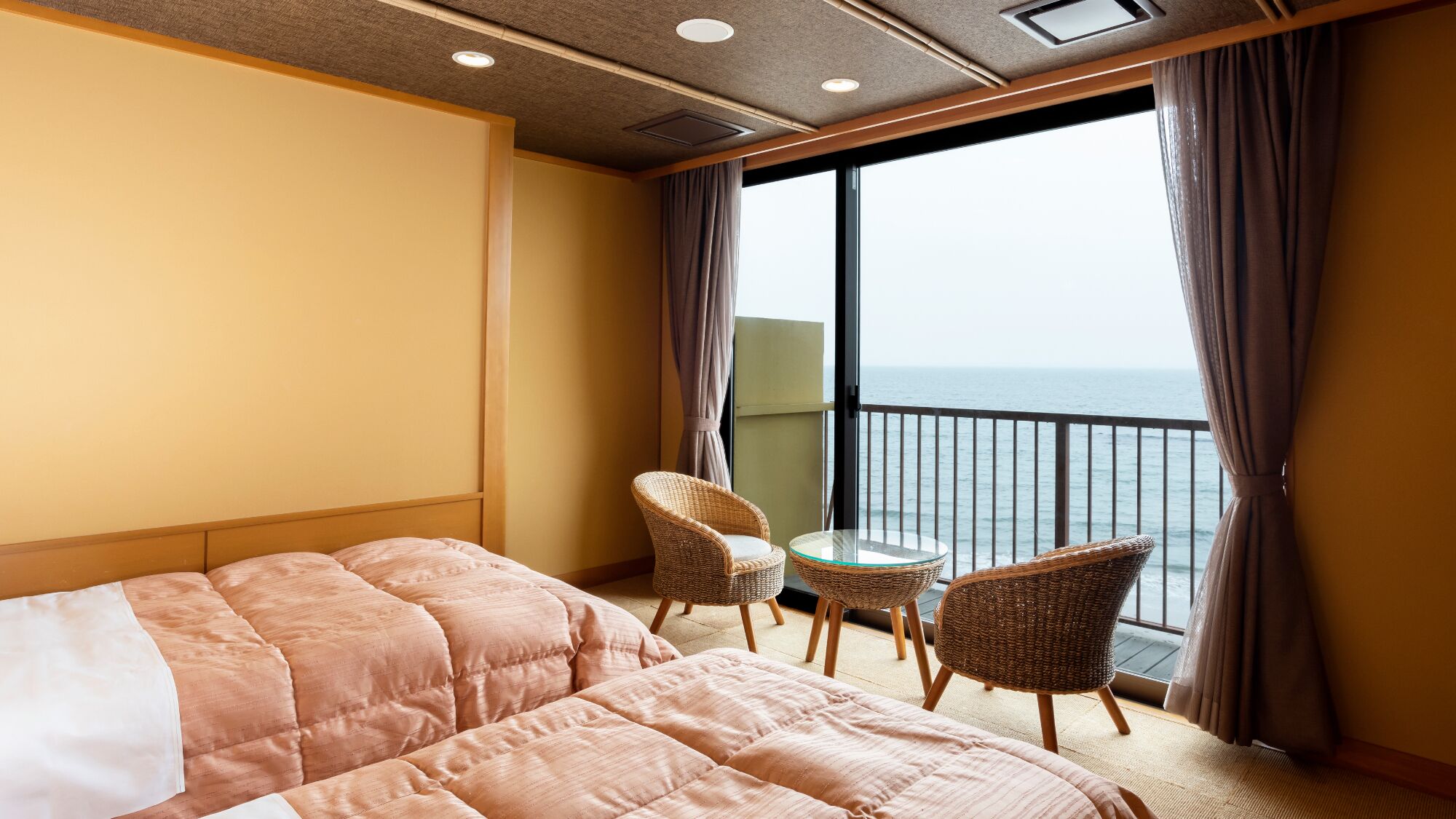 Quarto de hotel com uma cama, duas cadeiras de vime, uma mesinha e varanda com vista para o mar.