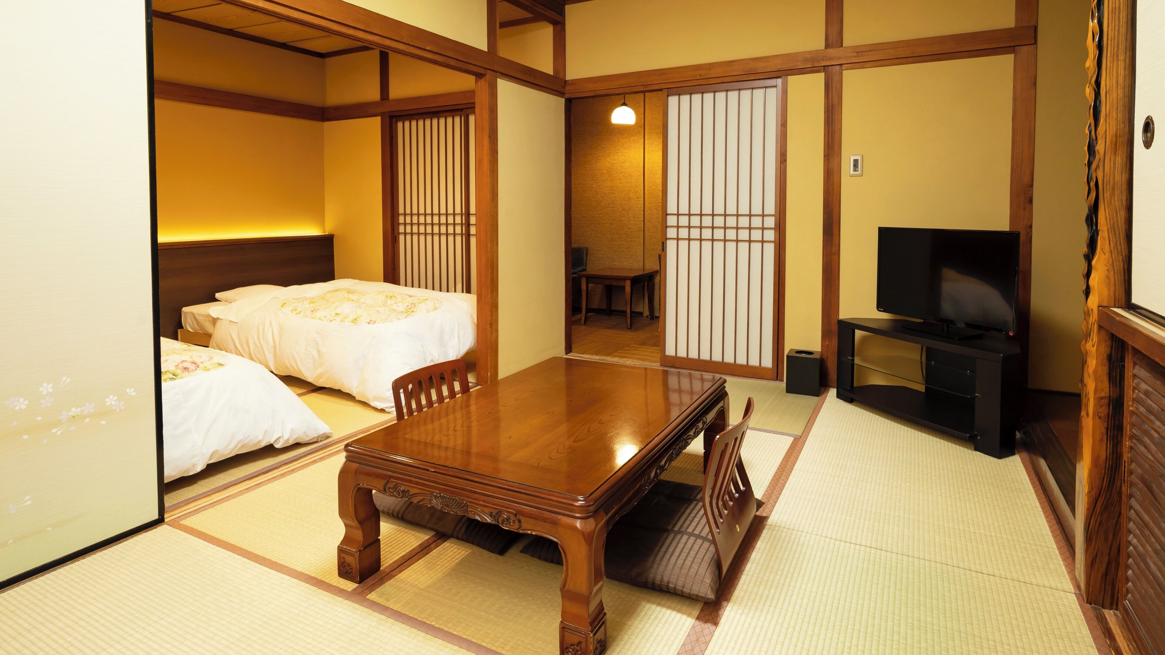 Um quarto japonês tradicional com tatames, uma mesa de madeira, duas camas, uma televisão e portas deslizantes.