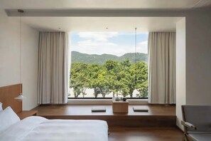 Quarto de hotel moderno com uma cama grande, uma mesa de cabeceira de madeira, uma mesinha e uma cadeira. O quarto possui uma janela ampla com cortinas, oferecendo vista para uma área arborizada e montanhas.