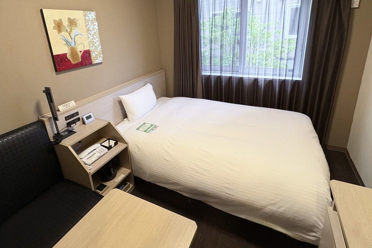 Quarto de hotel com cama, mesa de cabeceira com abajur, pequena escrivaninha com telefone e um quadro na parede.