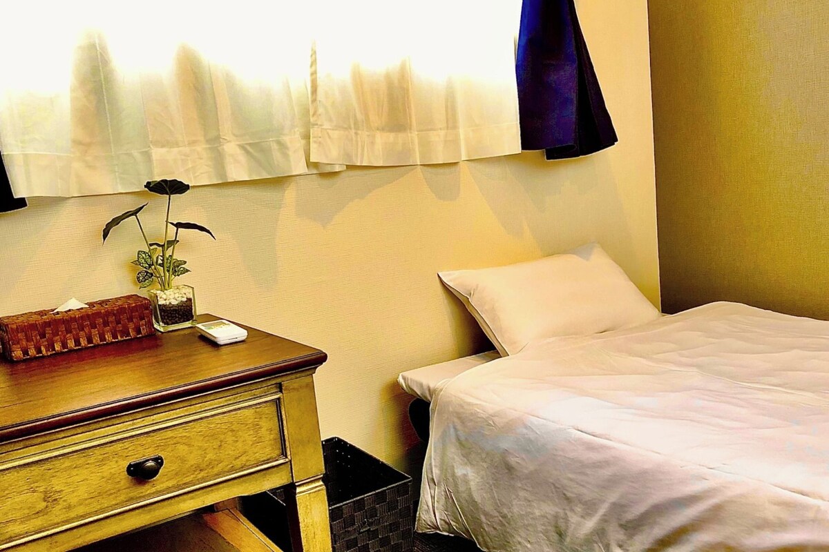 Quarto de hotel com uma cama, uma mesinha de cabeceira com abajur, uma pequena planta e uma janela com cortinas.