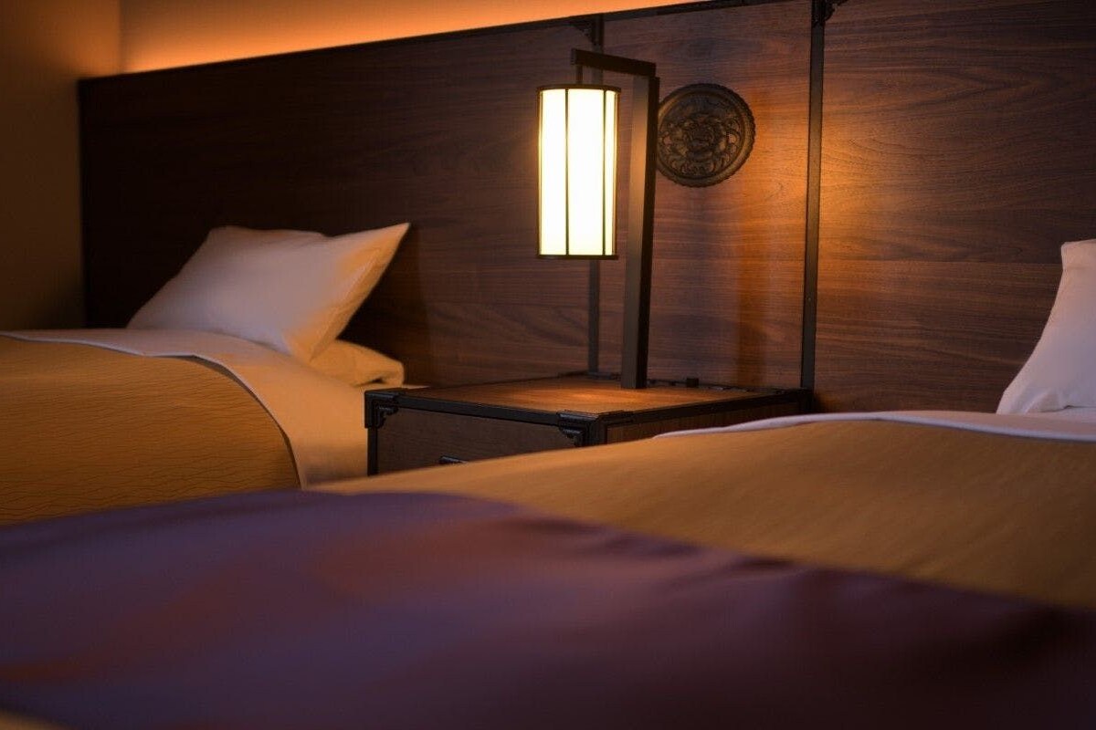 Quarto de hotel com duas camas, uma mesinha de cabeceira, uma luminária fixada na parede e um prato decorativo.