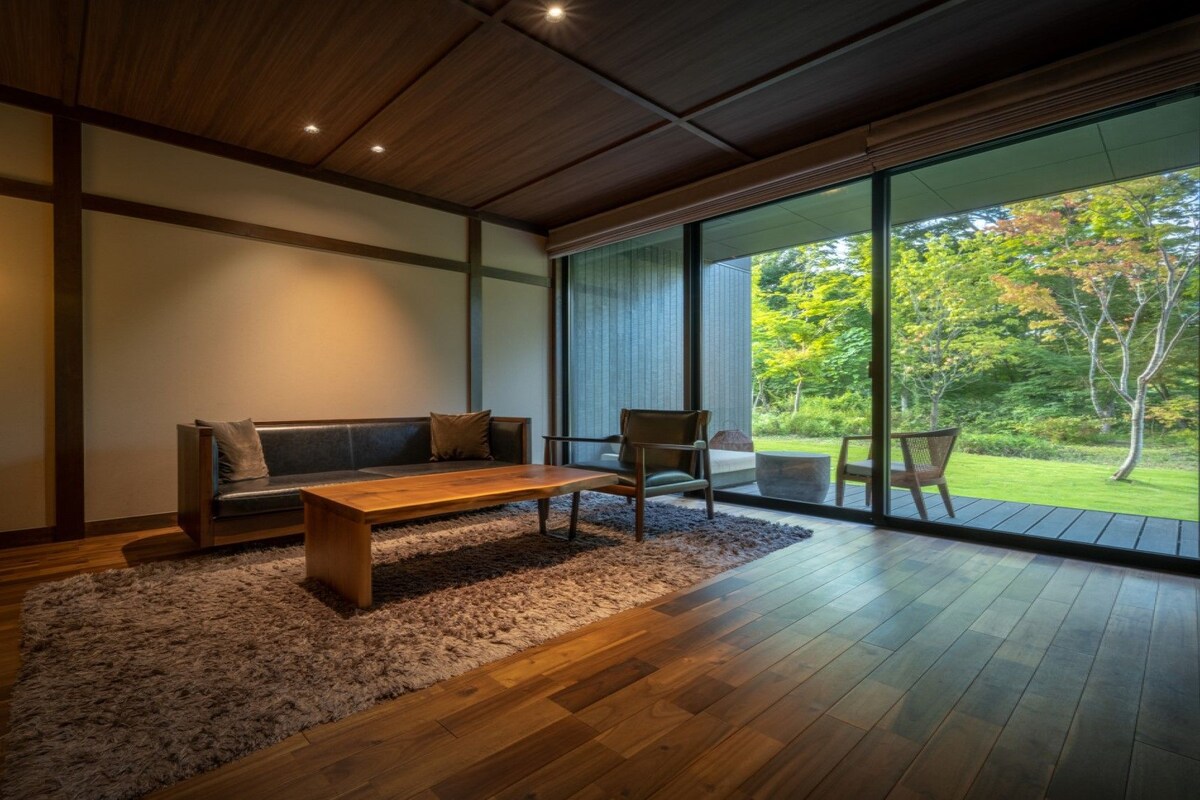 Uma sala de estar moderna com piso de madeira, um sofá, uma mesa de centro e uma janela grande que oferece vista para um jardim.