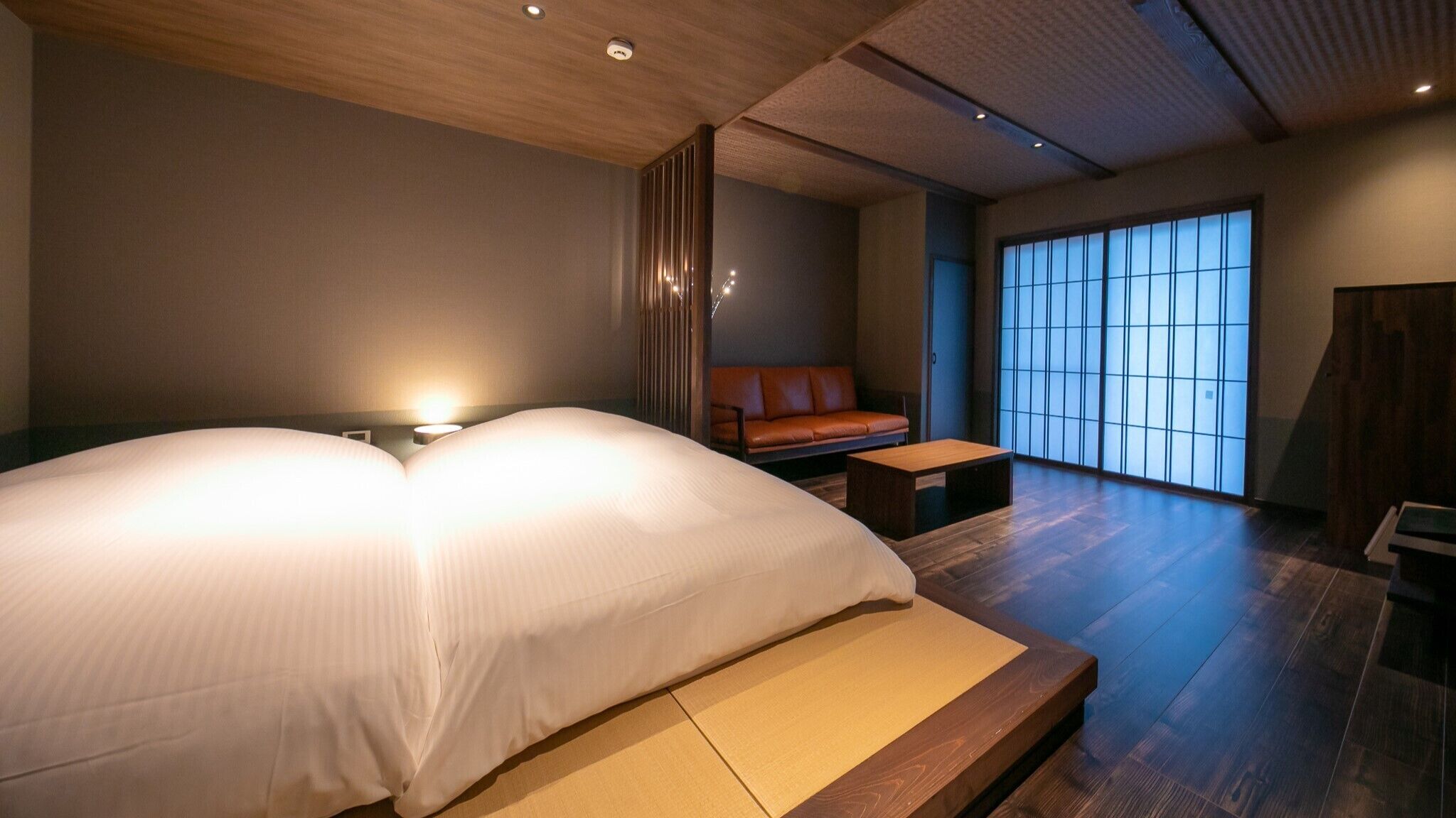 Quarto de hotel moderno com uma cama grande, um sofá pequeno, piso de madeira e uma porta de vidro deslizante.
