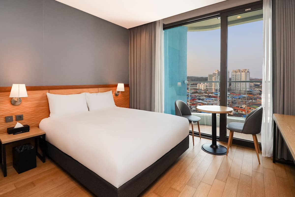 Quarto de hotel com uma cama grande, duas luminárias de cabeceira, uma mesinha com uma cadeira e vista para a cidade através de uma janela ampla.