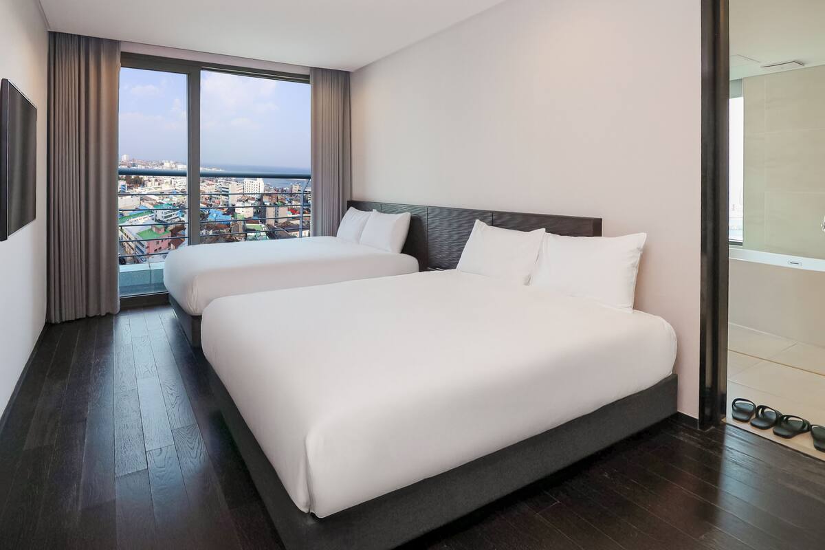 Quarto de hotel com duas camas, uma janela grande com vista para a cidade, piso de madeira escura e um espaço de banheiro pequeno.