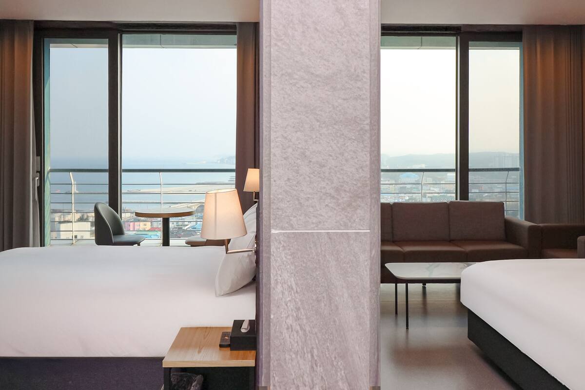 Quarto de hotel com uma cama grande, uma cama menor, um sofá, uma mesa e um abajur. Janelas amplas oferecem vista para a cidade e para um corpo d'água.