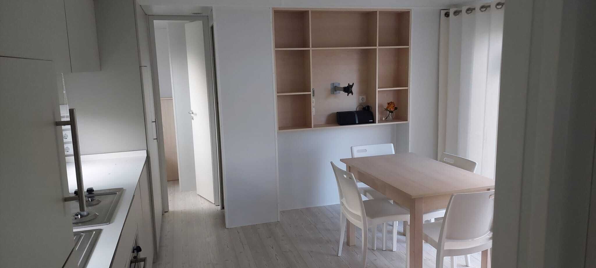 Cozinha moderna com armários brancos, uma mesa de jantar de madeira e cadeiras. Há uma prateleira embutida sobre a pia e uma porta que dá para outro cômodo.