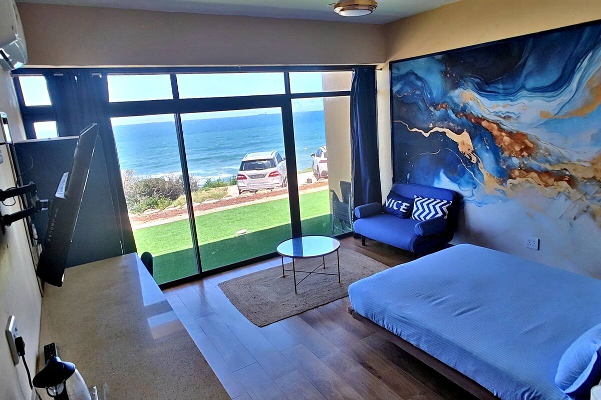 Quarto com cama, cadeira, mesinha e uma grande janela com vista para o mar.