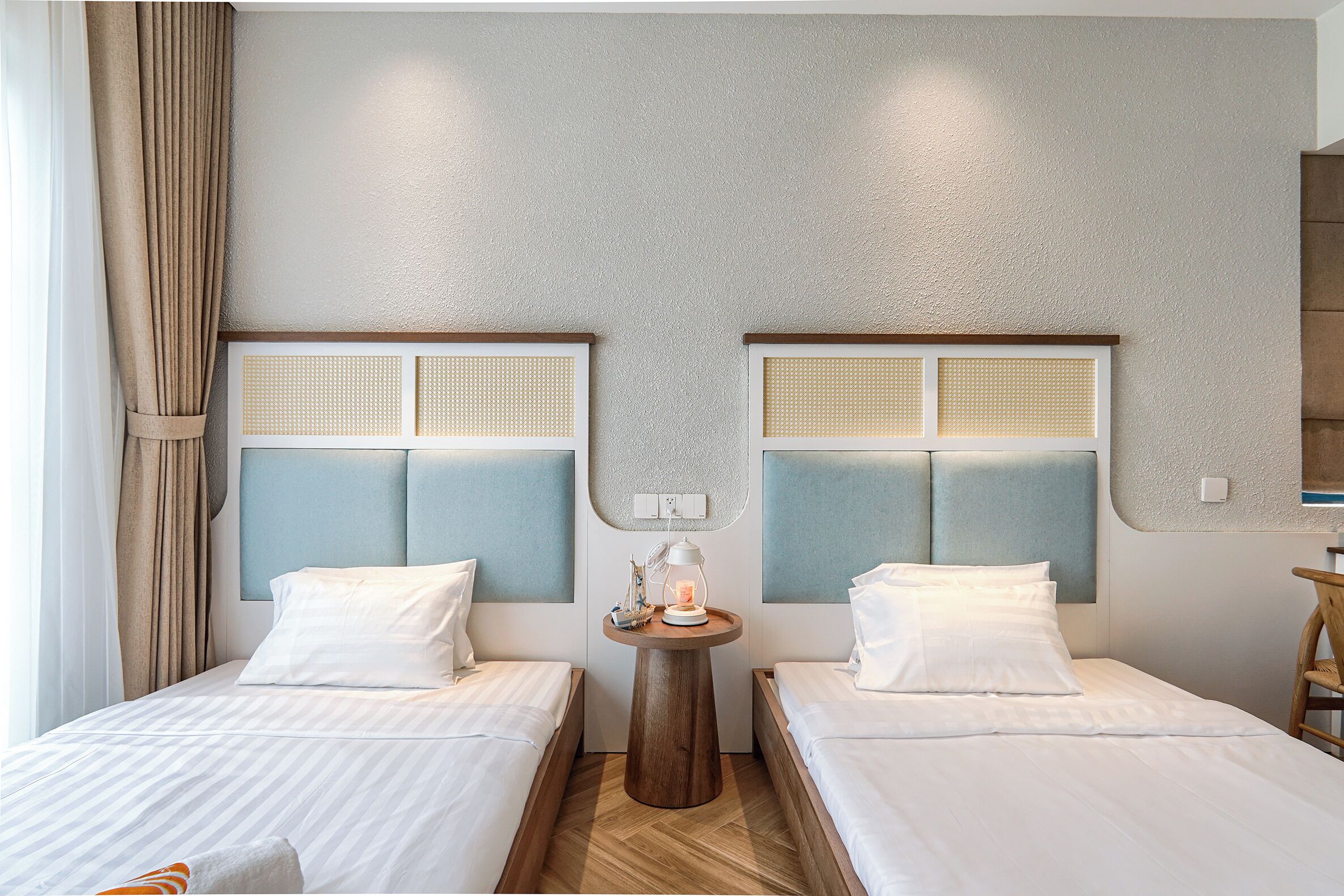 Quarto de hotel com duas camas, uma mesa lateral redonda de madeira e um abajur, além de uma parede com textura.