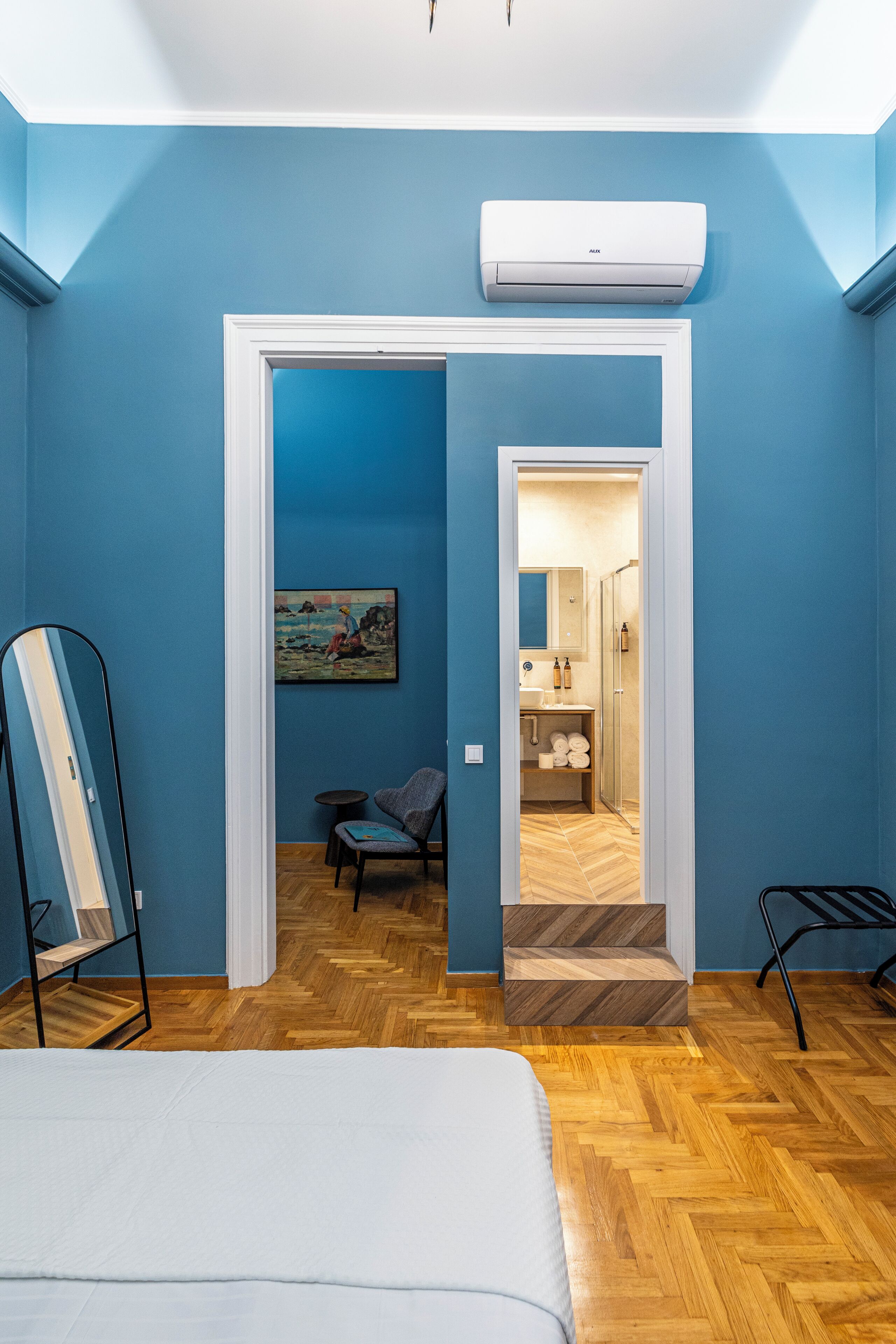 Quarto com cama, piso de madeira, parede azul, ar condicionado, quadro, cadeira e mesa pequena.