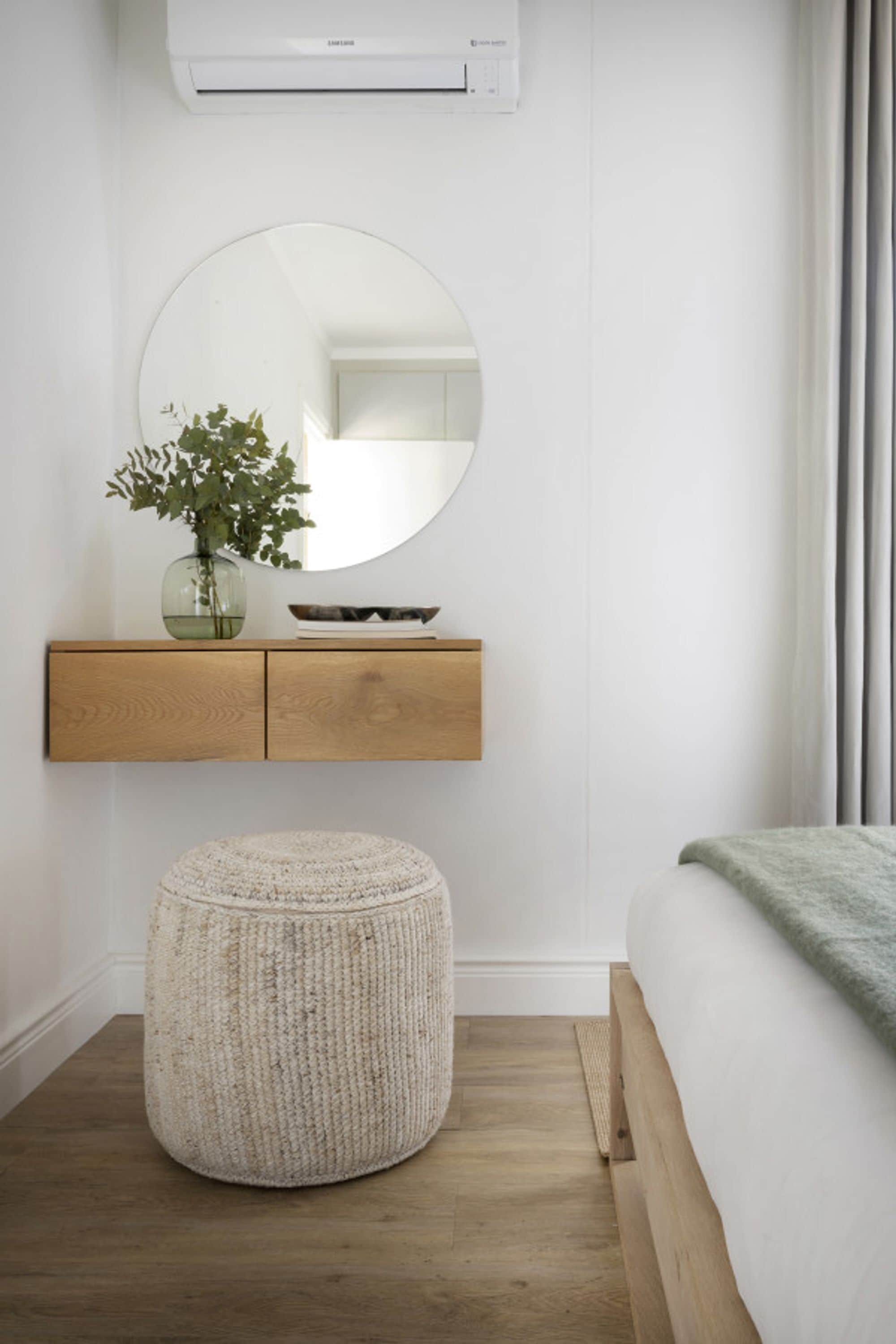 Quarto com cama de madeira, espelho redondo, otomano branco pequeno e mesa de cabeceira de madeira com um vaso e livros.