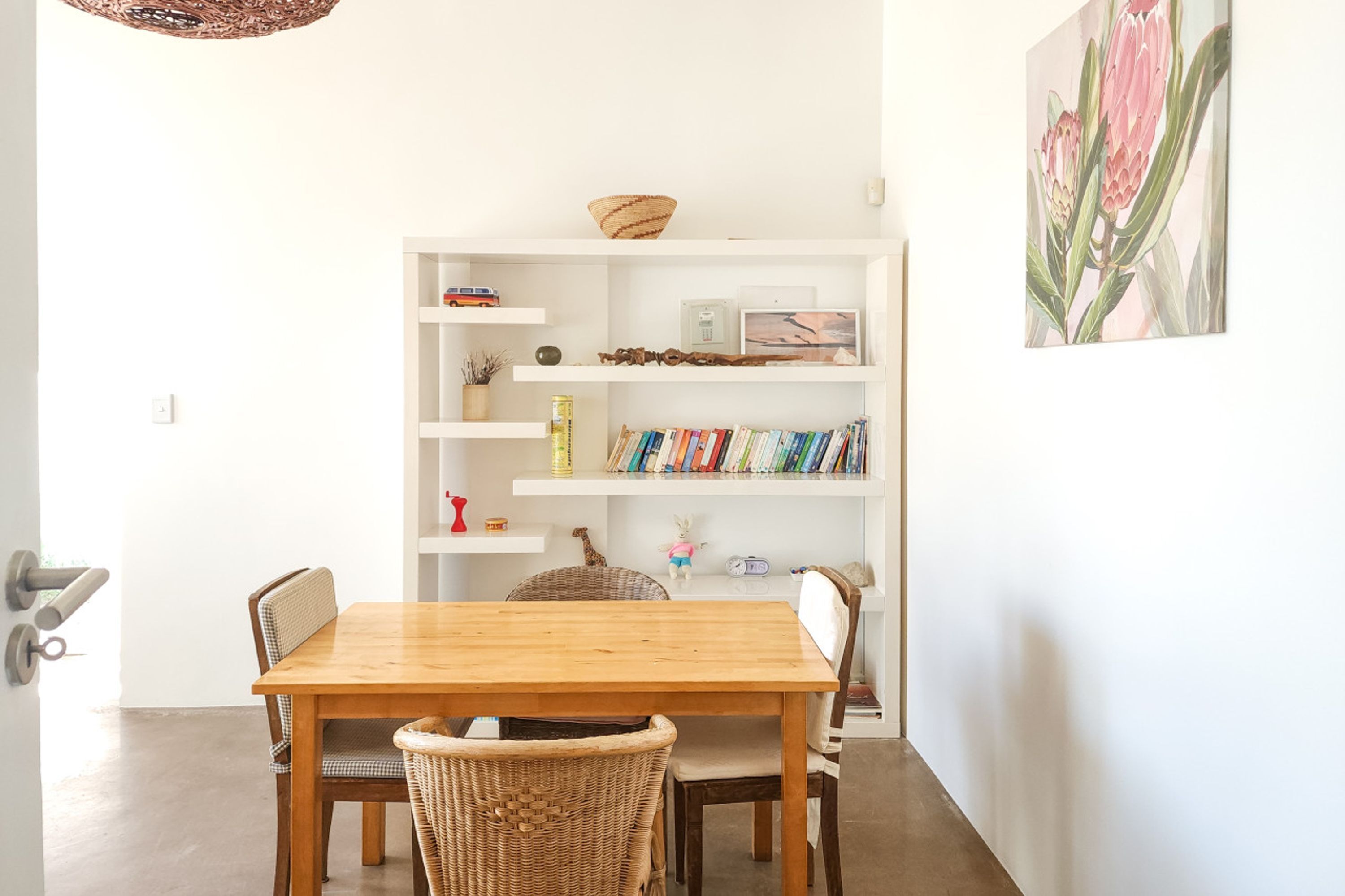 Uma sala de jantar com mesa de madeira e cadeiras de vime, uma estante branca com diversos objetos e um quadro de flores na parede.