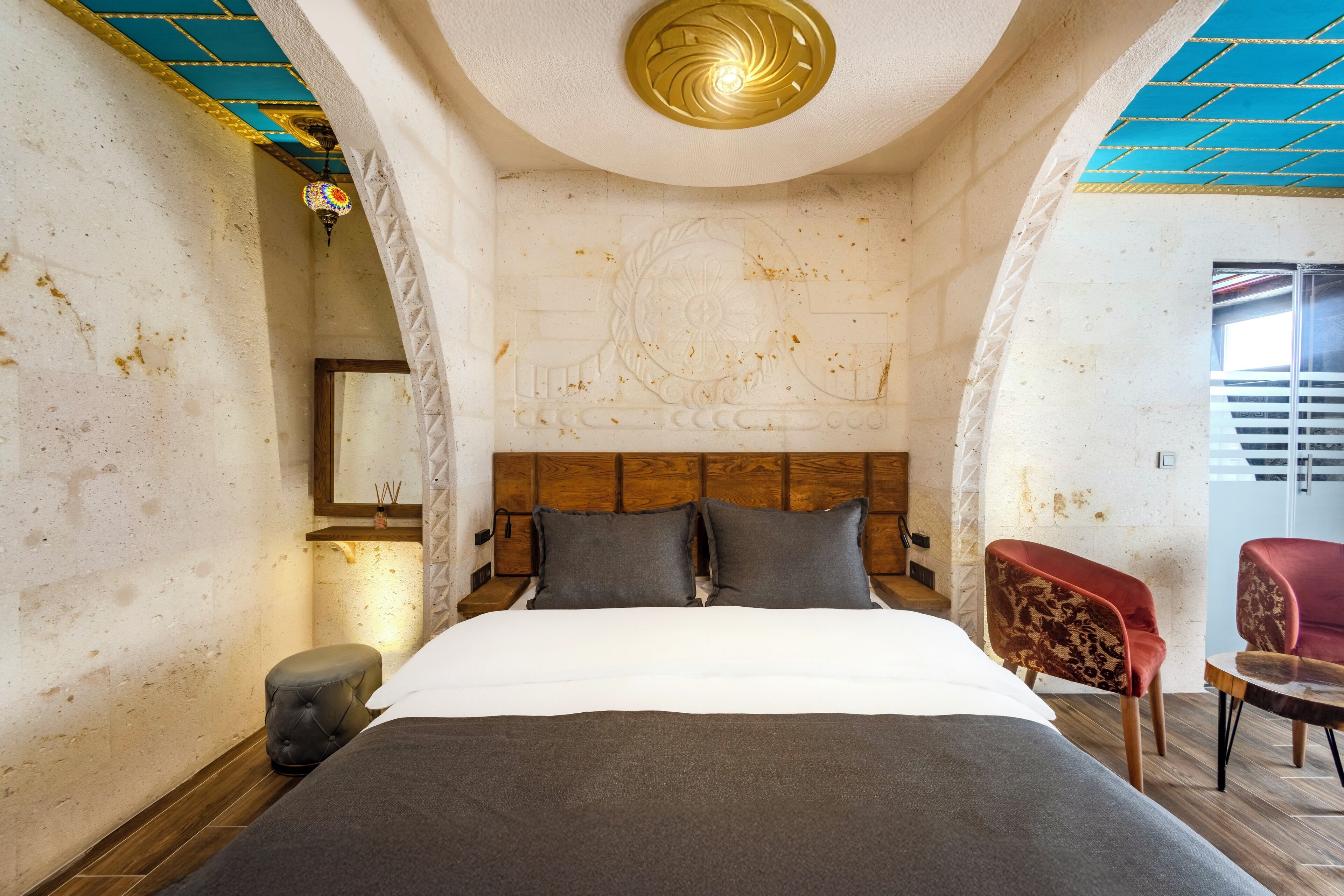 Quarto com cama grande, cabeceira de madeira, um banquinho pequeno e duas cadeiras.