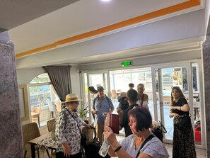 Um grupo de pessoas em um saguão com entrada por porta de vidro, uma mesa e cadeiras.