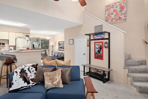 Uma sala de estar com um sofá azul, um banco de madeira, uma escada e uma cozinha ao fundo.