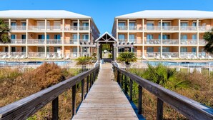 Um resort com uma passarela de madeira que leva a uma área de piscina, cercada por palmeiras e um céu azul límpido.