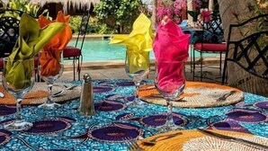 Uma área de refeições à beira da piscina, com decoração tropical, incluindo palmeiras e uma estrutura de telhado de palha.