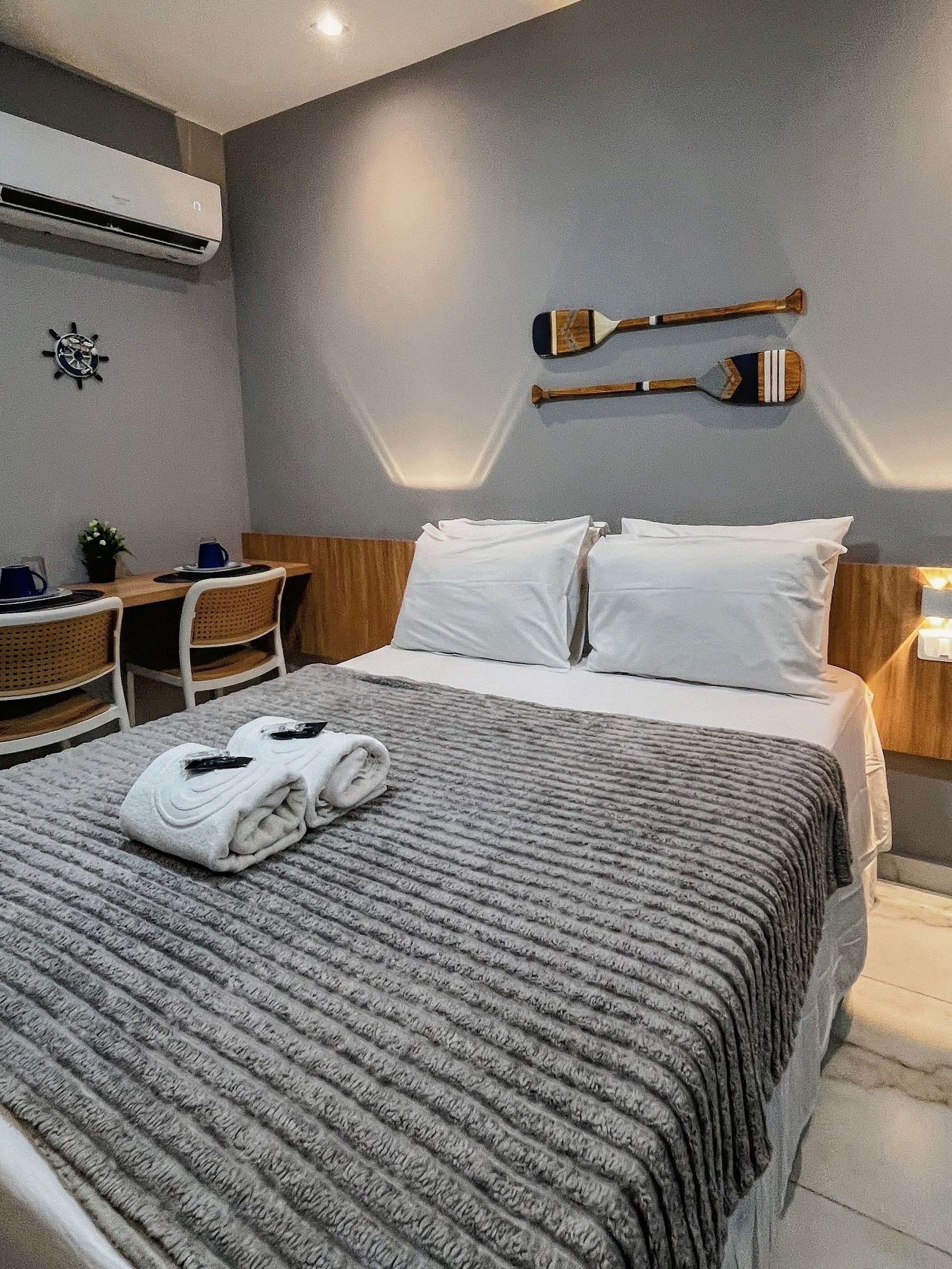 Quarto de hotel com cama, escrivaninha e uma cadeira. Há um ar condicionado fixado na parede e um objeto decorativo.
