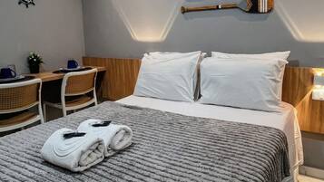 Quarto de hotel com cama, escrivaninha e uma cadeira. Há um ar condicionado fixado na parede e um objeto decorativo.