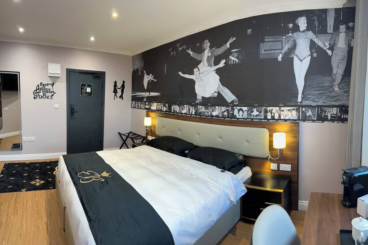 Quarto de hotel com cama, escrivaninha, cadeira, máquina de café e parede com obras de arte com tema de dança.