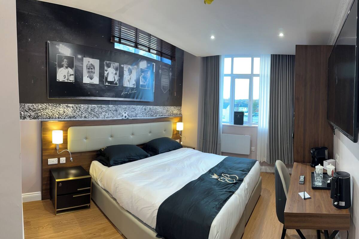 Quarto de hotel com uma cama grande, mesas de cabeceira, escrivaninha com cafeteira, televisão e uma parede com fotografias emolduradas.