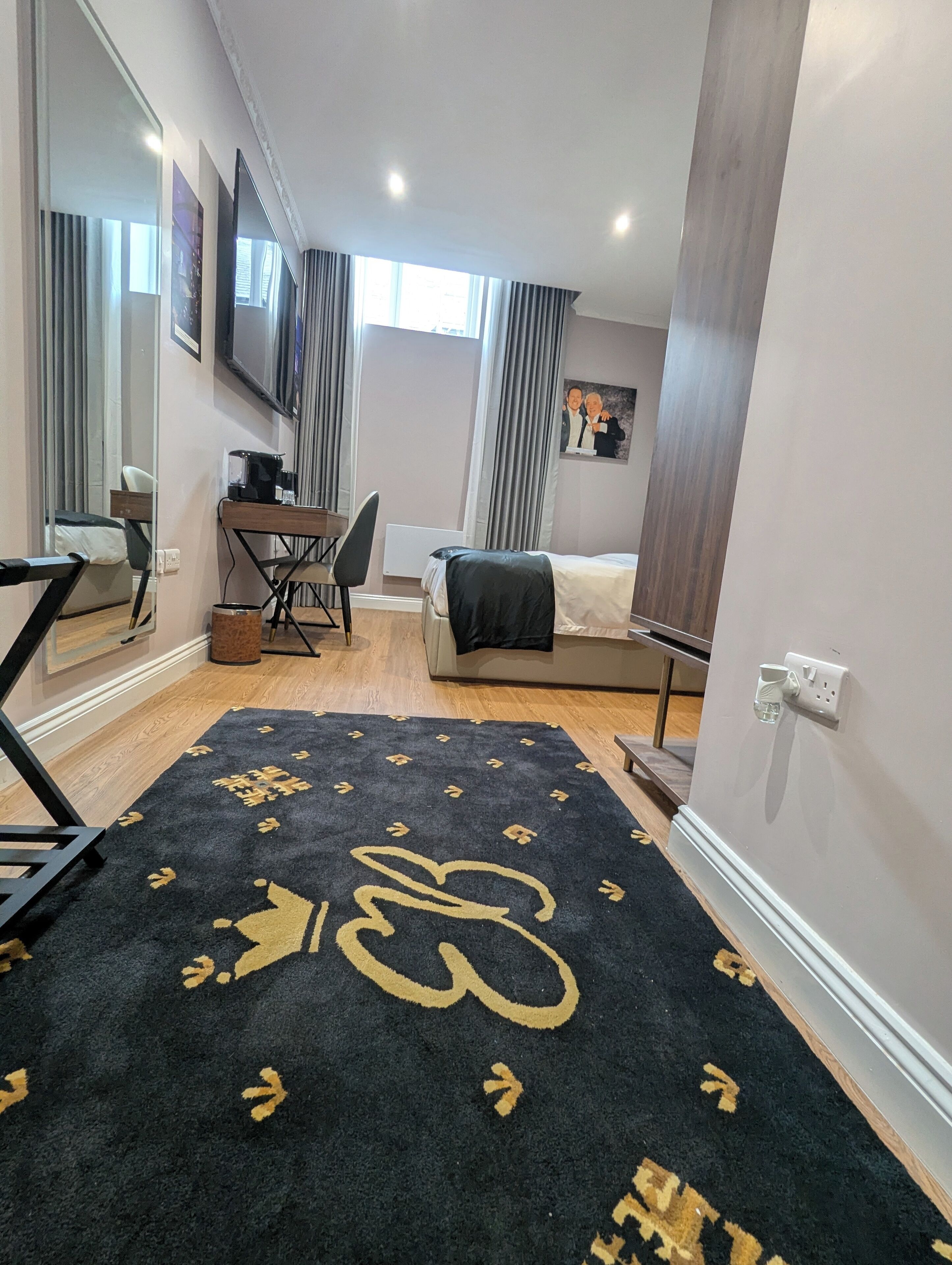Quarto de hotel com cama, escrivaninha, cadeira, máquina de café, televisão e um espelho grande.