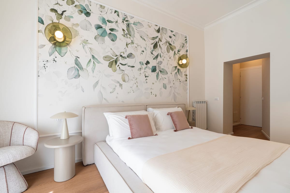 Quarto com cama, mesa de cabeceira, abajur e parede com padrão floral.