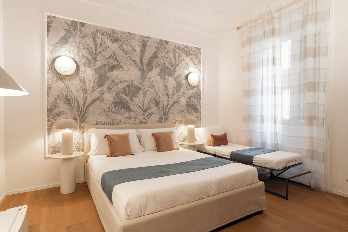 Quarto de hotel com duas camas, uma tapeçaria grande na parede e uma janela com cortinas leves.