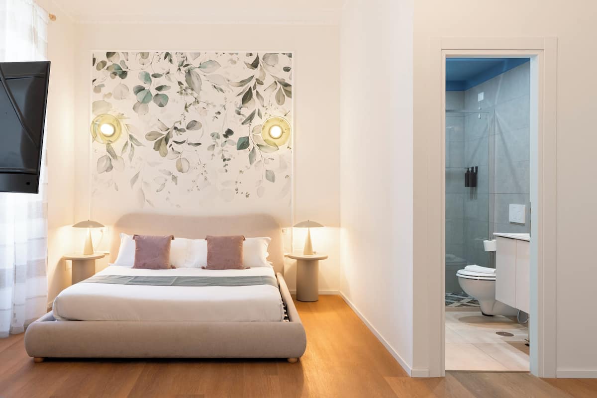 Quarto com cama, duas luminárias de cabeceira, TV fixada na parede e banheiro visível por uma porta aberta.