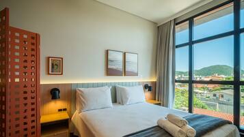 Quarto de hotel com uma cama, duas luminárias fixadas na parede, uma janela com vista para um horizonte urbano e um separador vermelho com padrão quadriculado.