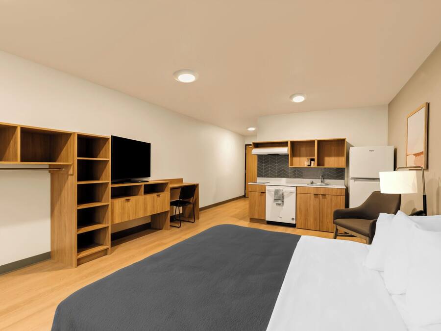 Quarto de hotel moderno com uma cama grande, uma escrivaninha com televisão de tela plana, uma cozinha compacta e um espaço de estar com uma poltrona.