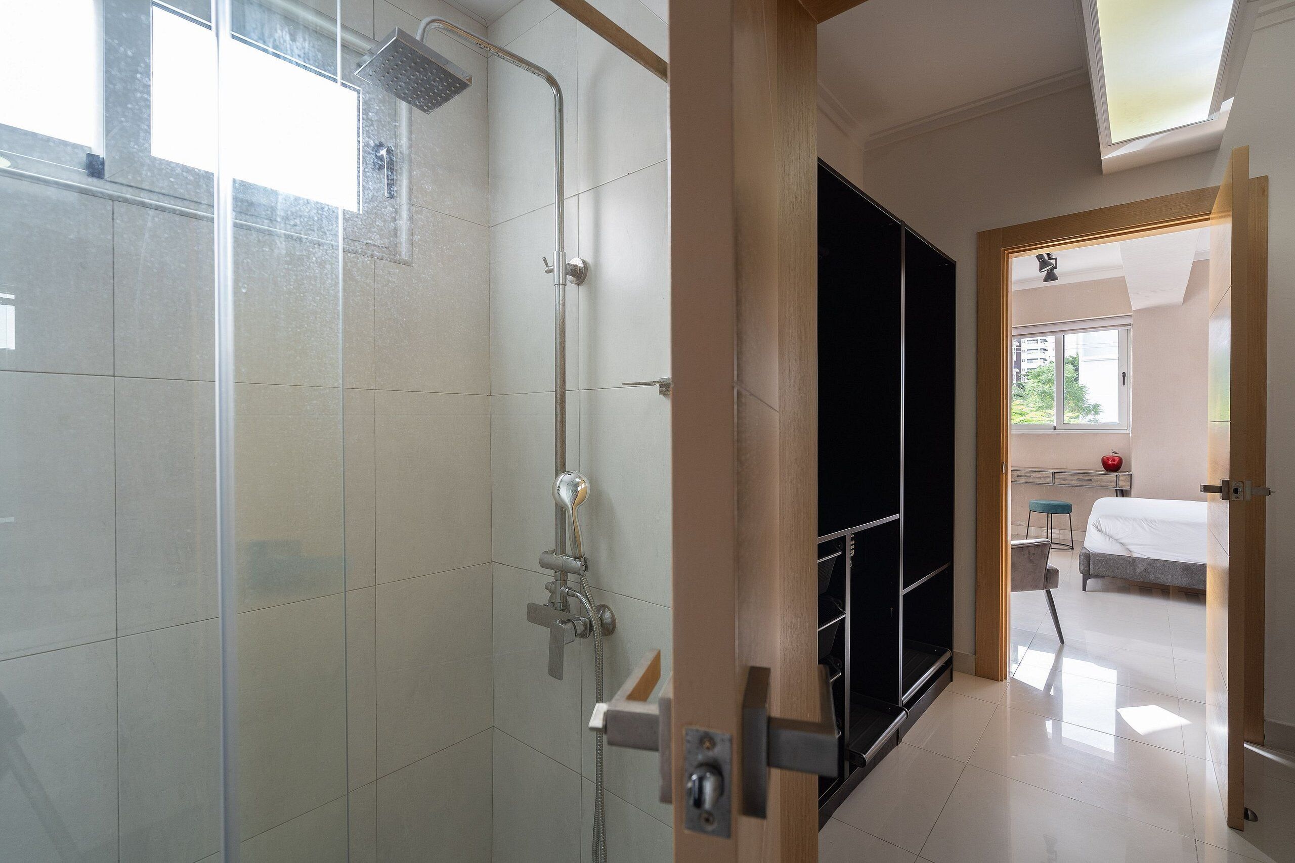 Uma casa de banho moderna com cabine de chuveiro em vidro, sanita visível e porta que dá para um quarto com cama e poltrona.
