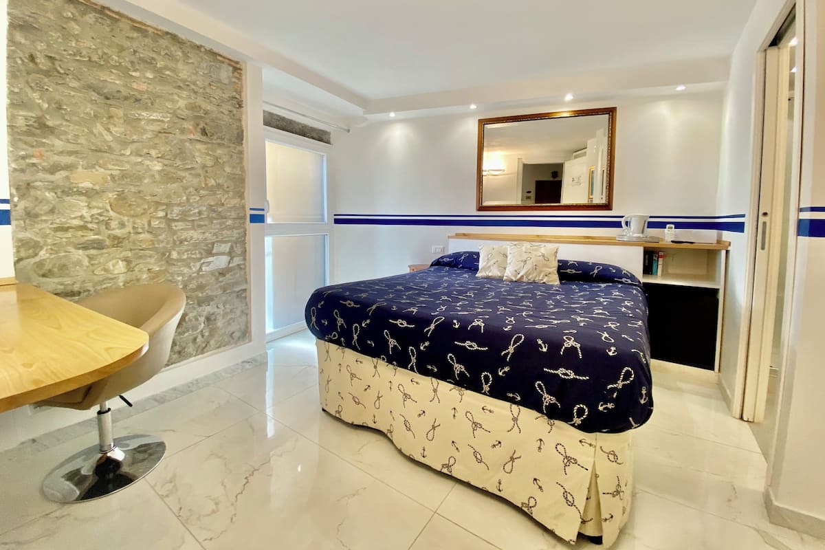 Quarto de hotel com cama, escrivaninha, cadeira, parede de pedra, espelho e prateleira com objetos.