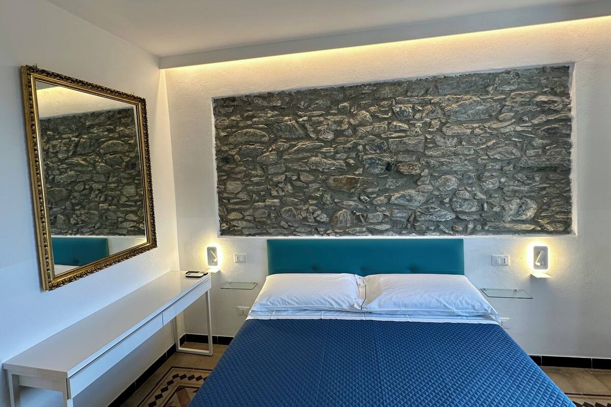 Quarto com parede de pedra, cama com roupa de cama branca e azul, mesa de cabeceira branca e espelho com moldura dourada.