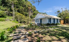 Uma casa térrea com fachada branca e detalhes azuis, cercada por um jardim bem cuidado e um muro de pedra.