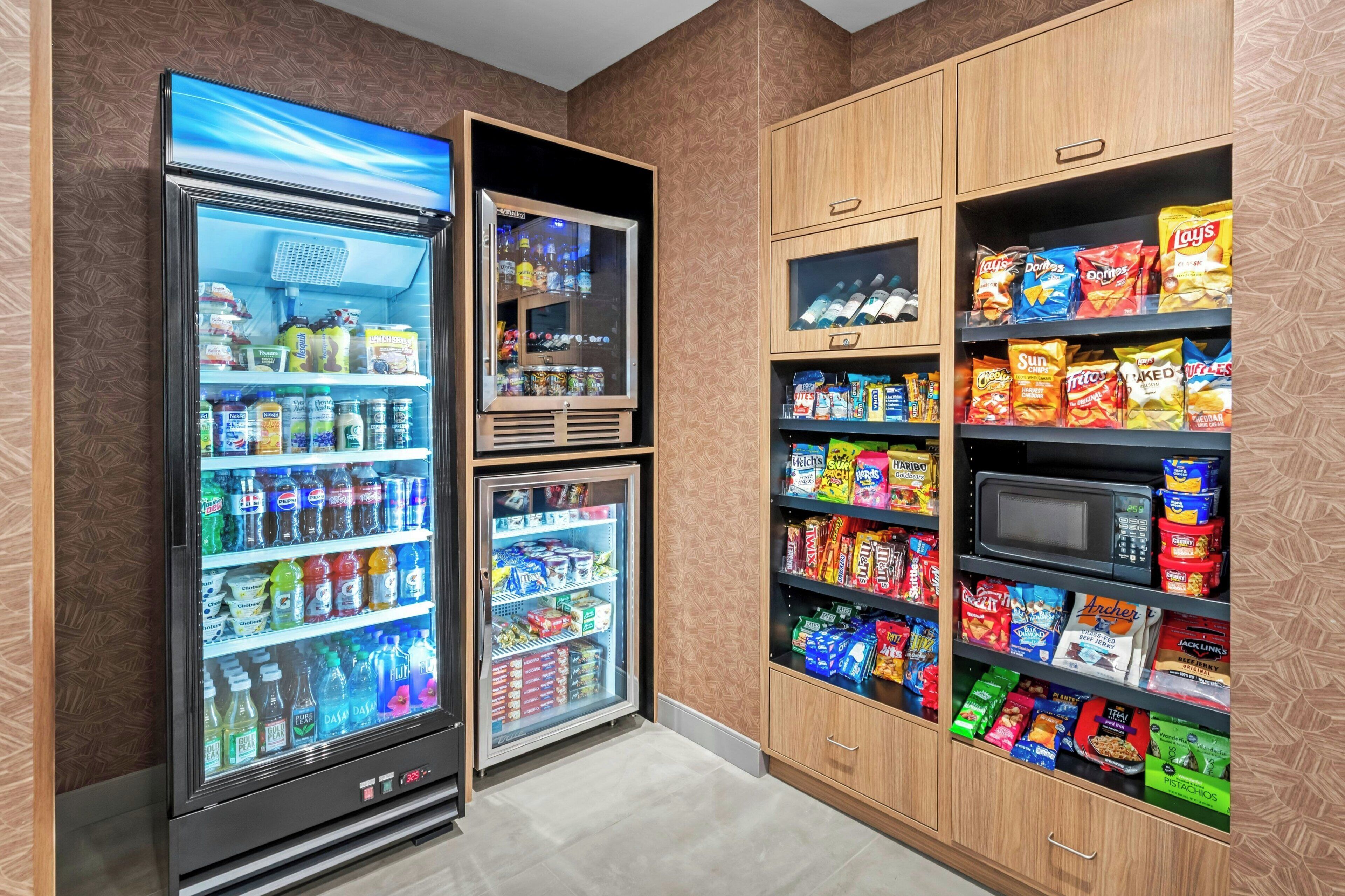 Uma área de venda automática com frigorífico, micro-ondas e estantes repletas de snacks e bebidas.
