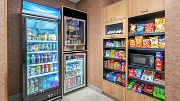 Uma área de venda automática com frigorífico, micro-ondas e estantes repletas de snacks e bebidas.