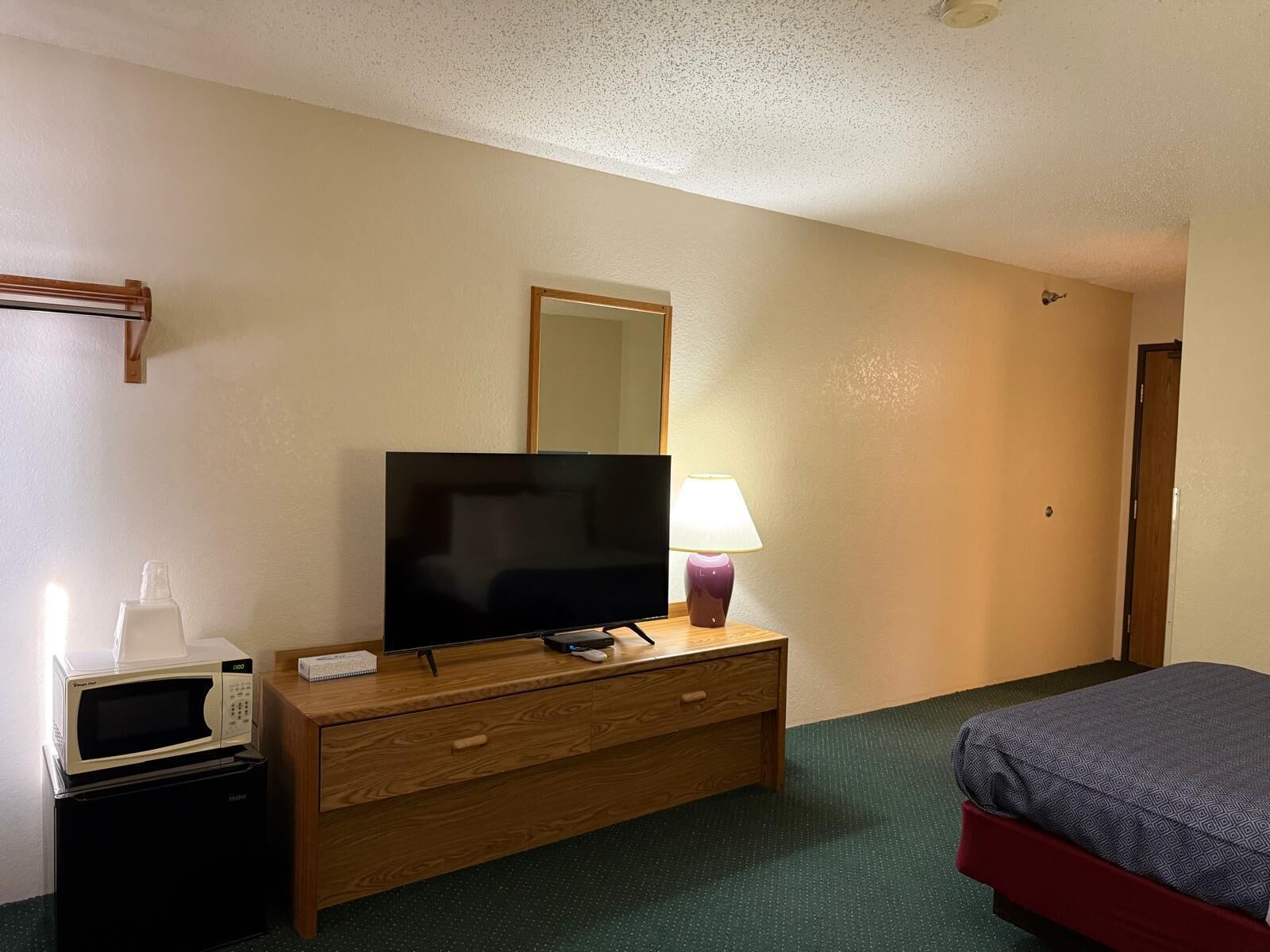 Quarto de hotel com cómoda de madeira, televisão de ecrã plano, micro-ondas, candeeiro e cama com colcha estampada.