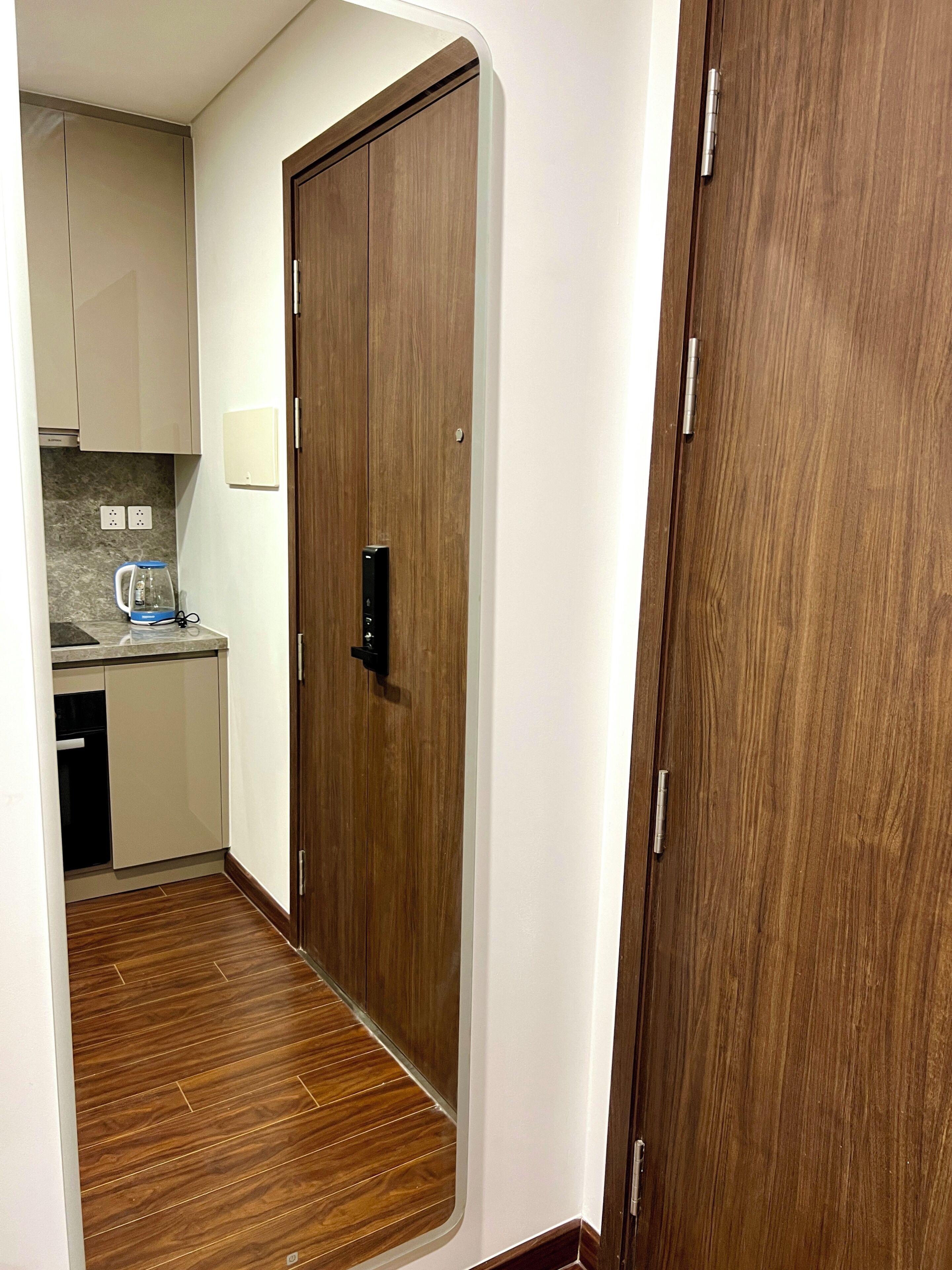 Um corredor com porta de madeira, área de cozinha com armários, cafeteira e tomada.