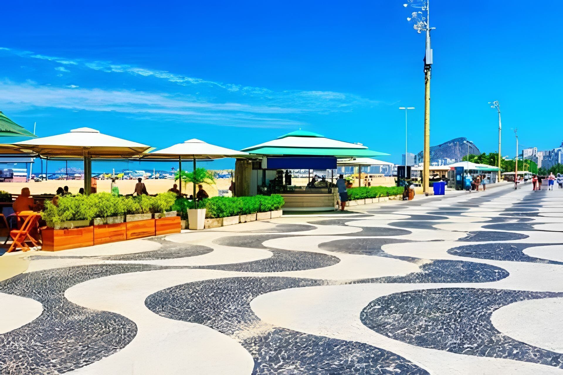 Um calçadão à beira-mar com guarda-sóis, áreas de estar e vista para a praia e montanhas.