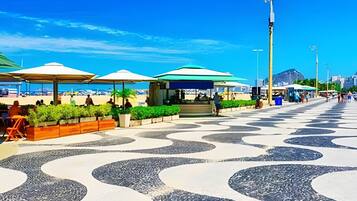 Um calçadão à beira-mar com guarda-sóis, áreas de estar e vista para a praia e montanhas.