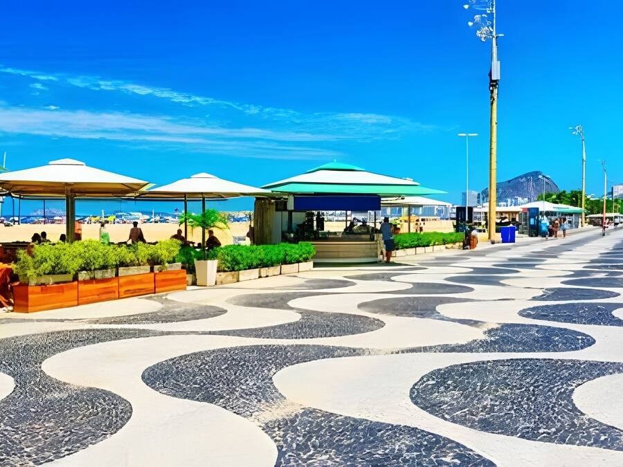 Um calçadão à beira-mar com guarda-sóis, áreas de estar e vista para a praia e montanhas.