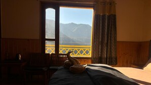 Quarto com cama, cadeira, mesa com chaleira e janela com vista para montanhas.
