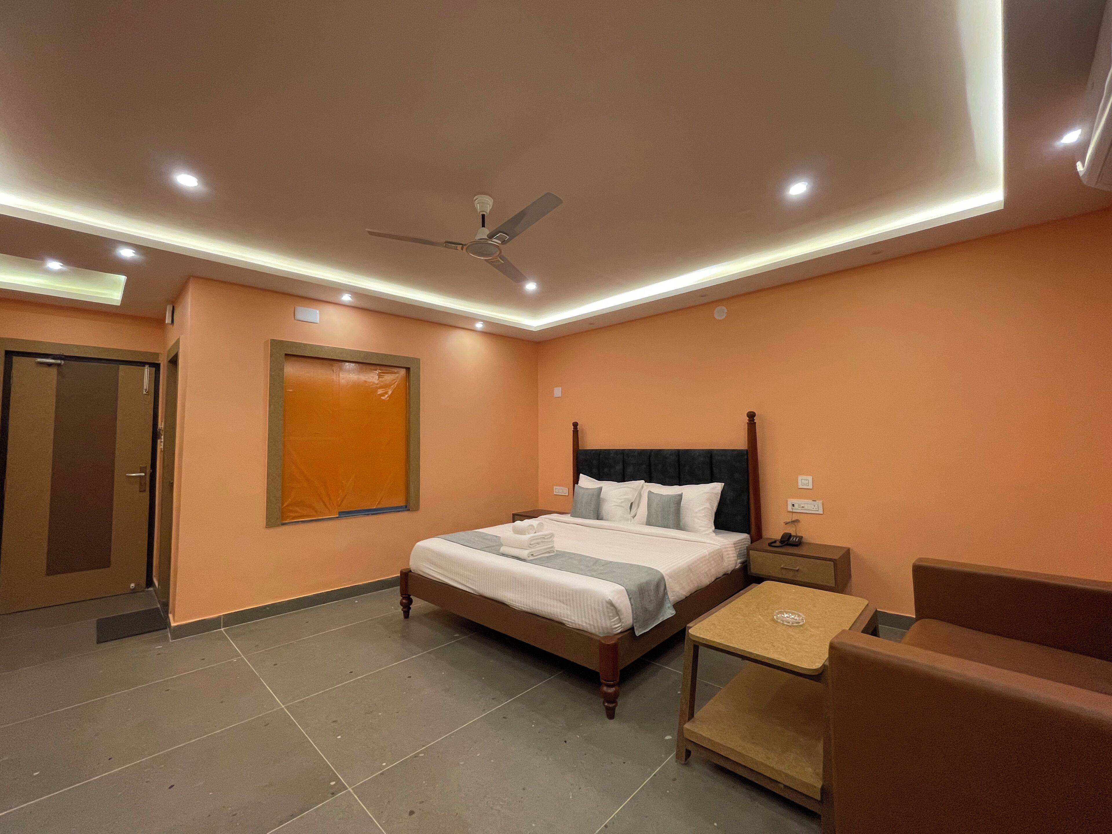 Quarto de hotel com cama, mesa de cabeceira, cadeira, ventoinha no teto e uma janela grande.
