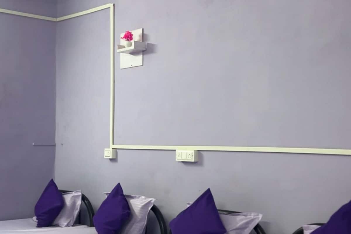 Quarto com duas camas, cada uma com colchão branco e almofadas roxas, uma prateleira pequena branca com uma flor rosa e uma tomada elétrica branca na parede.